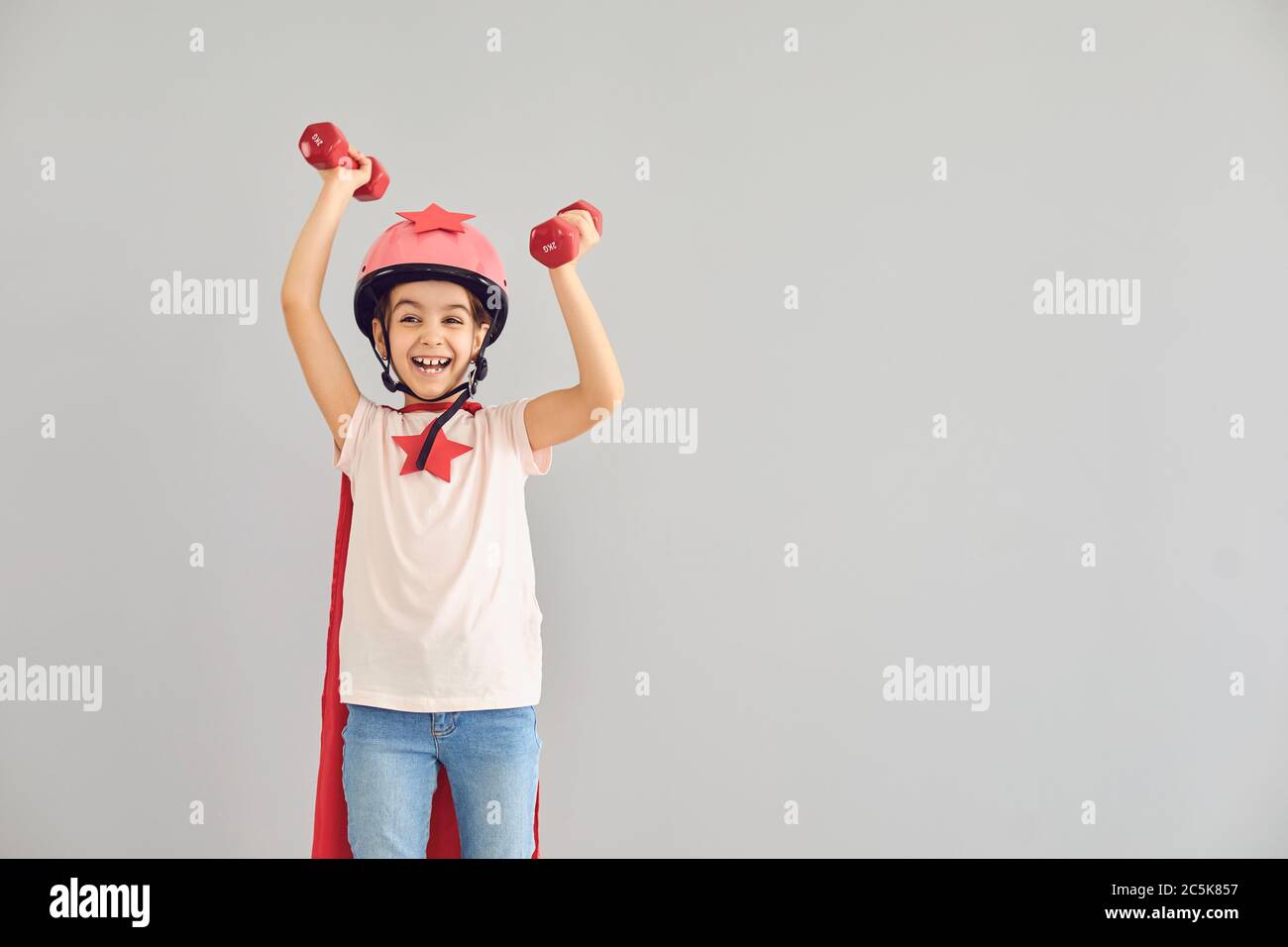 A child in a costume of a super hero raised his hands up with dumbbells ...