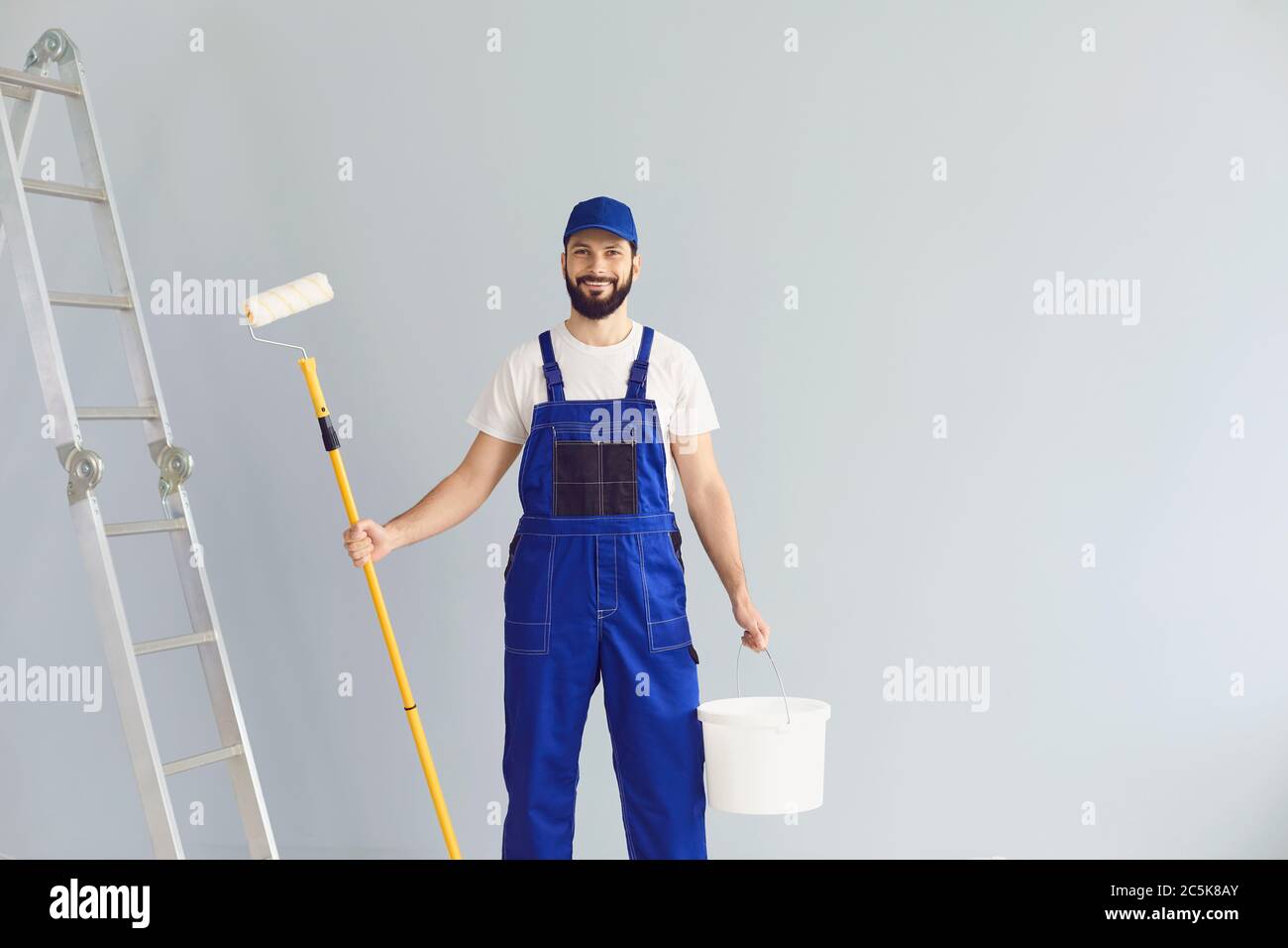 Repair worker in uniform with roller brush and bucket of paint on grey ...