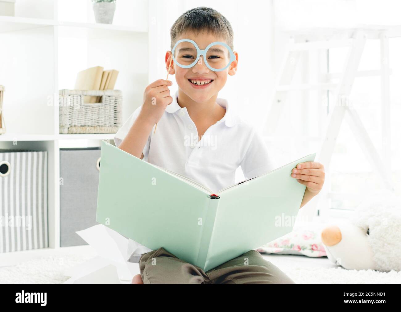 Cute little boy reading nice book wearing paper glasses in a kids room ...