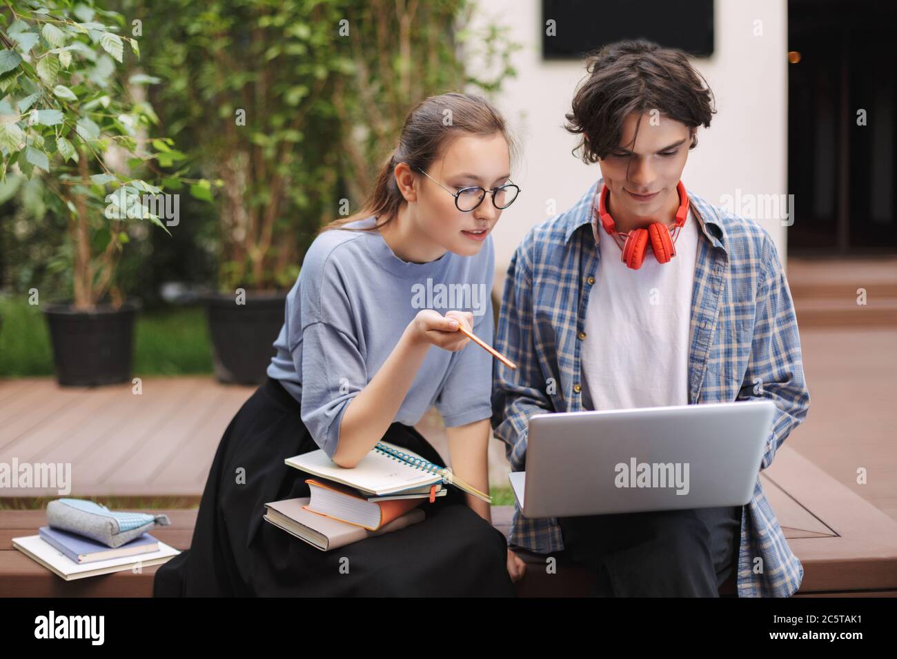 Photo of two students sitting on bench with books and working on laptop ...