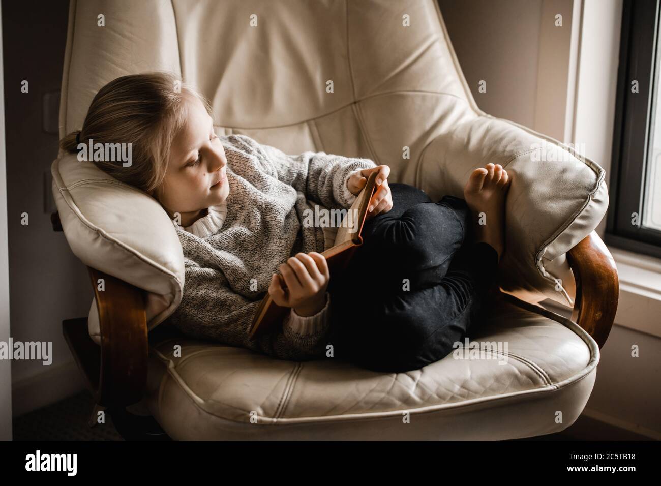 A blonde Caucasian girl sitting curled up on a chair reading a book ...