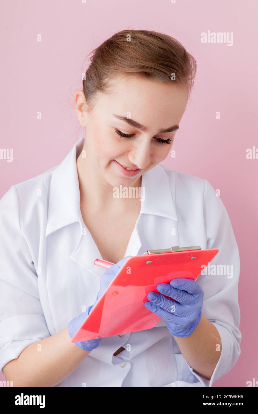Portrait of young female doctor making notes in notebook on pink ...