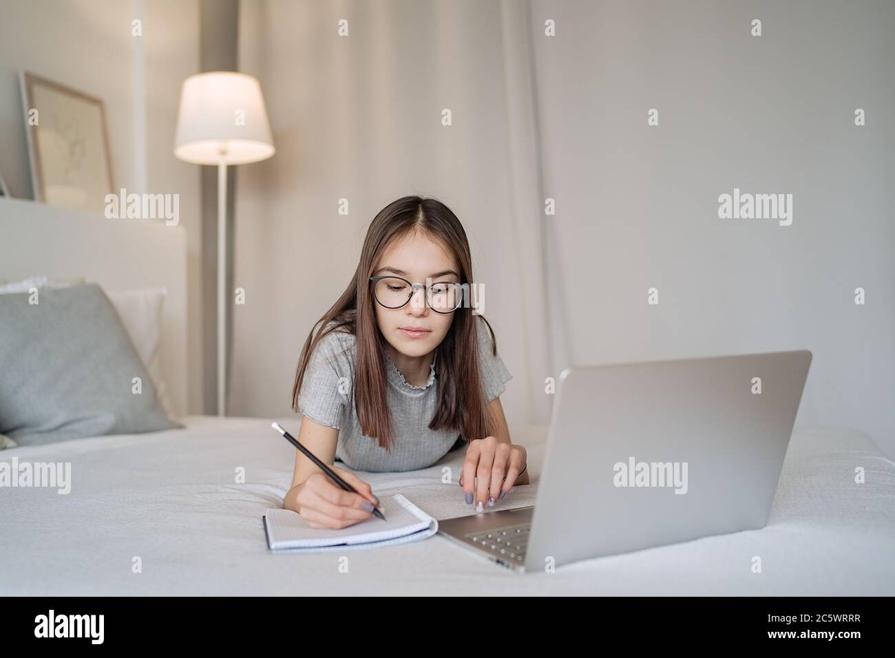 Teenager girl doing homework lying on bed at home. Student writing down ...