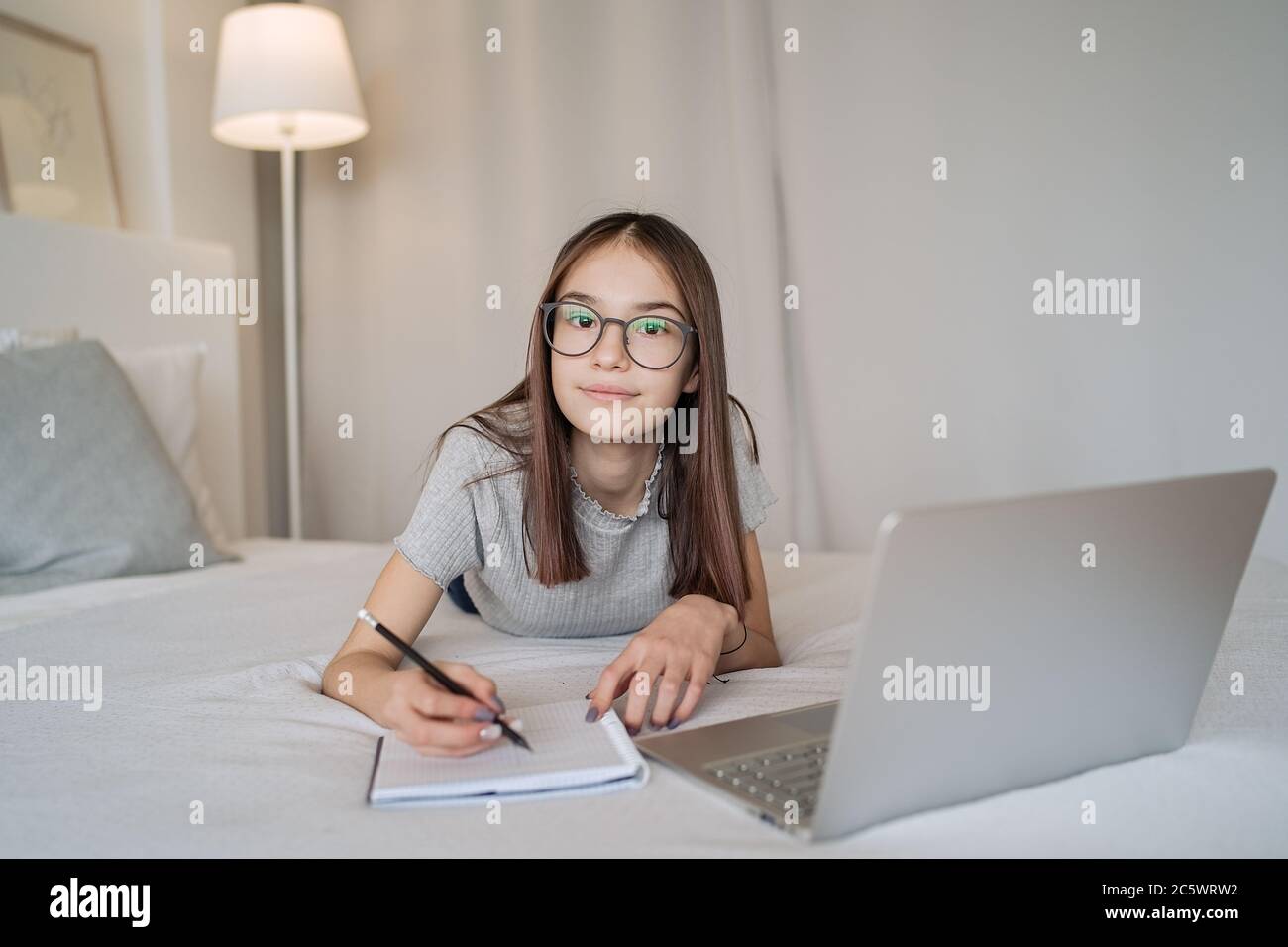 Teenager girl doing homework lying on bed at home. Student writing down ...