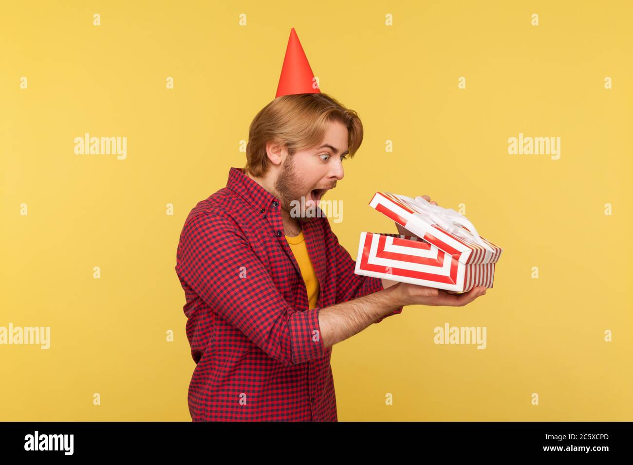 Astonished guy in checkered shirt and party cone opening gift box ...