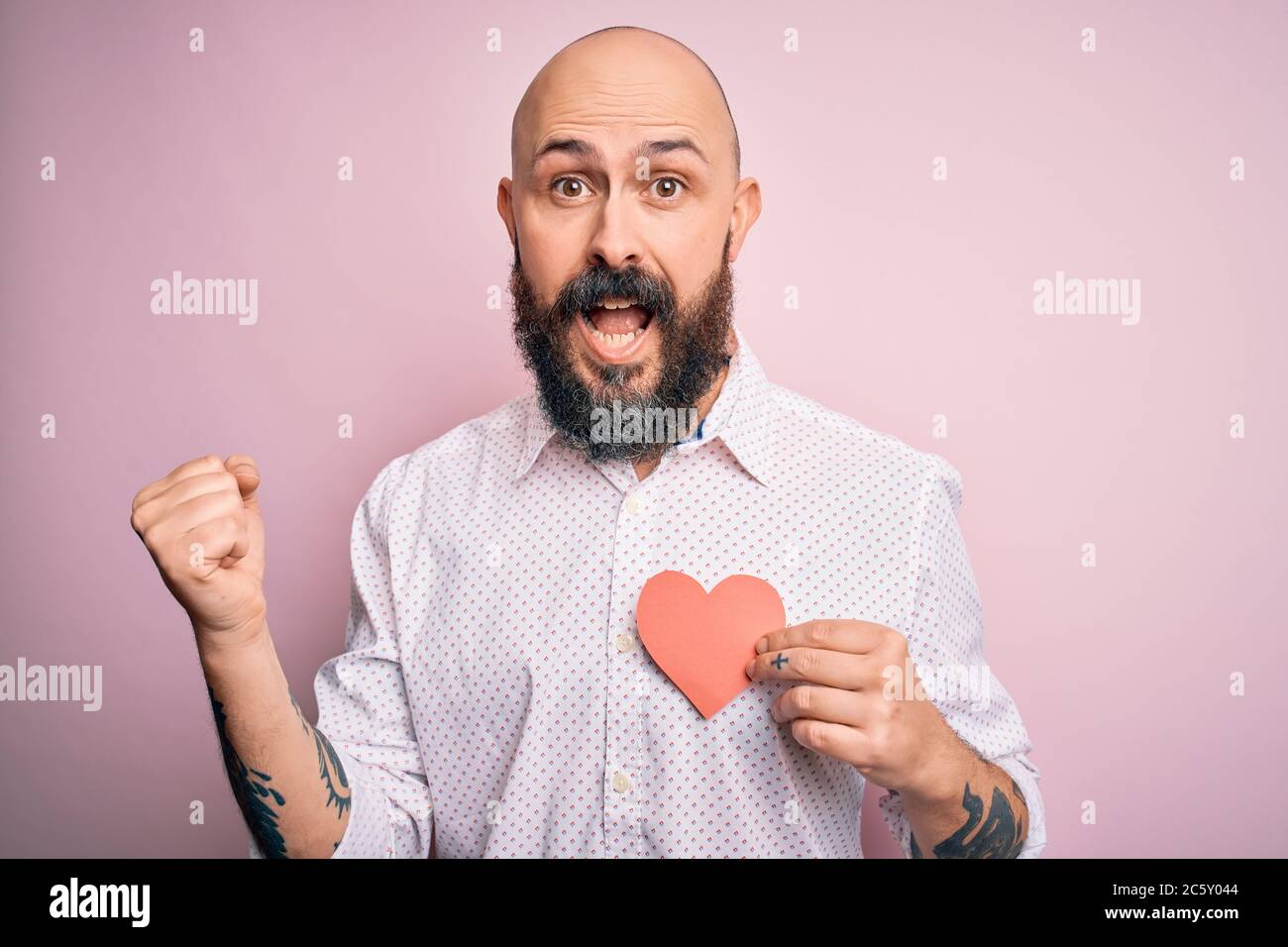 Handsome romantic bald man with beard holding red heart paper over pink ...