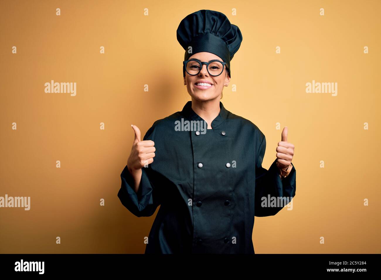 Young beautiful brunette chef woman wearing cooker uniform and hat over ...