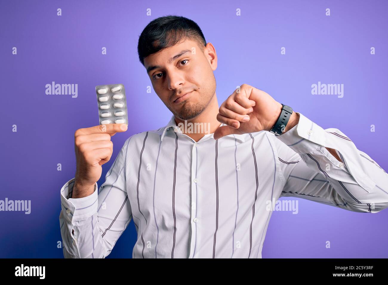 Young hispanic man holding pharmaceutical antibiotics pills over purple ...