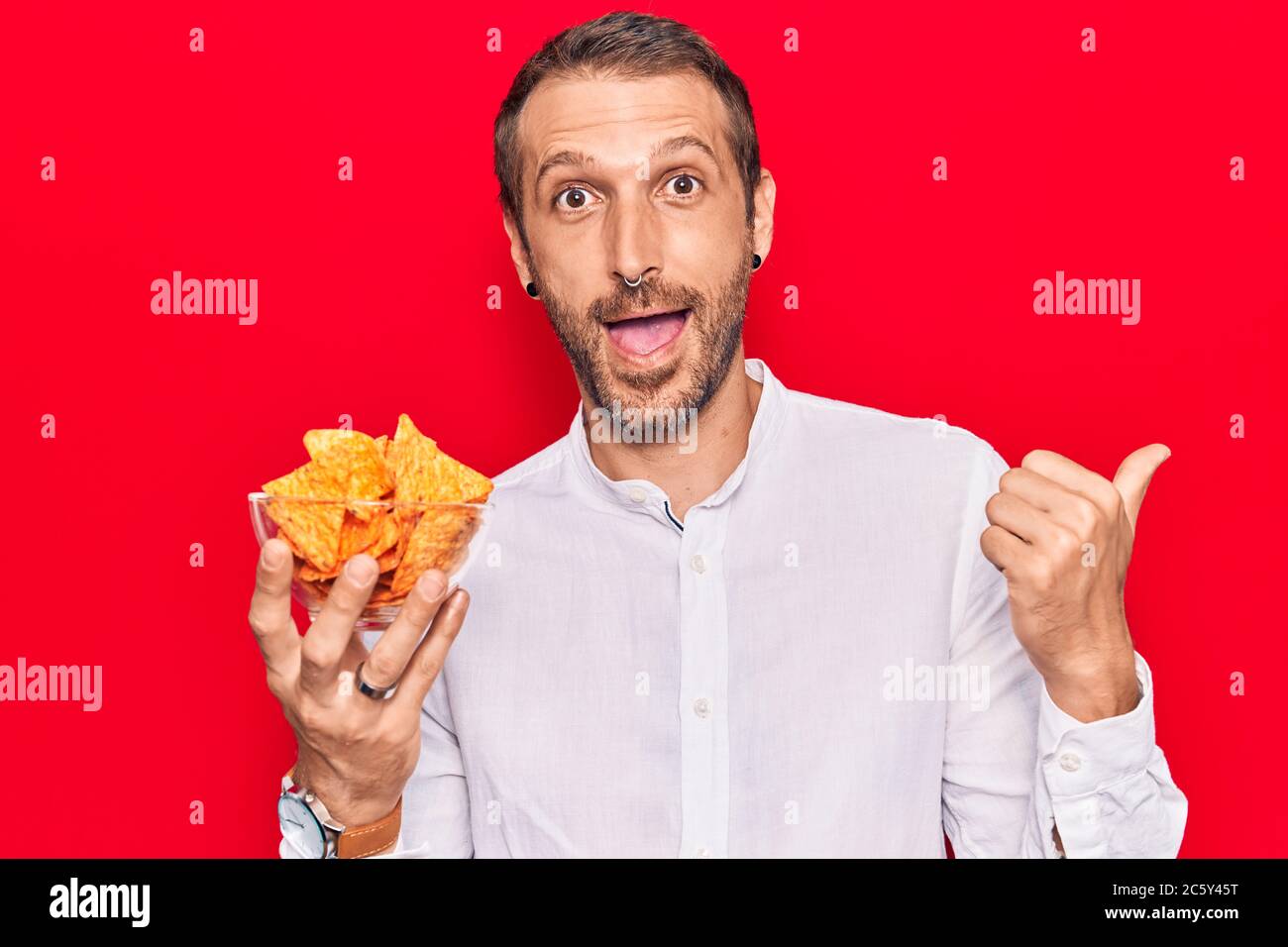 Young handsome man holding nachos potato chips pointing thumb up to the ...