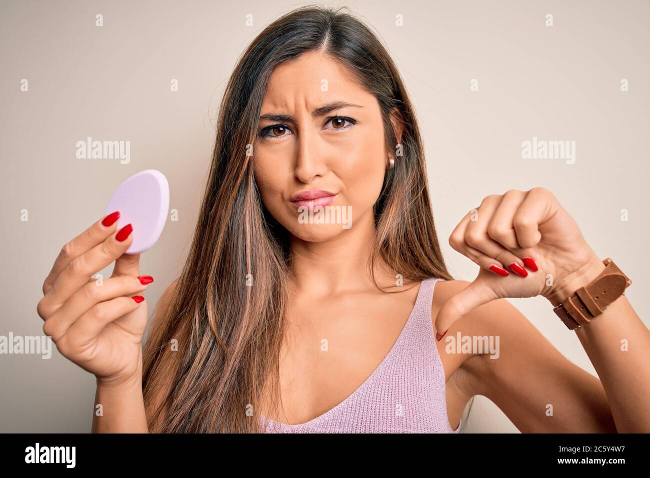 Young beautiful woman using beauty sponge with facial makeup over pink ...