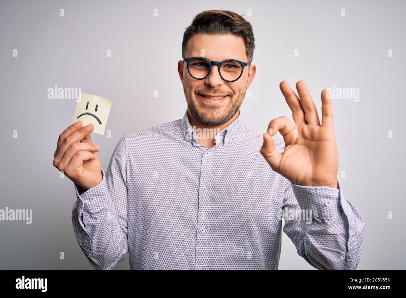 Young business man with blue eyes holding paper note with sad face as ...