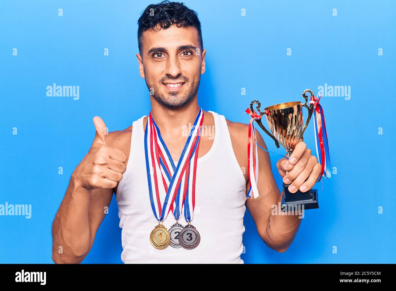 Young hispanic man wearing winner medals holding trophy smiling happy ...