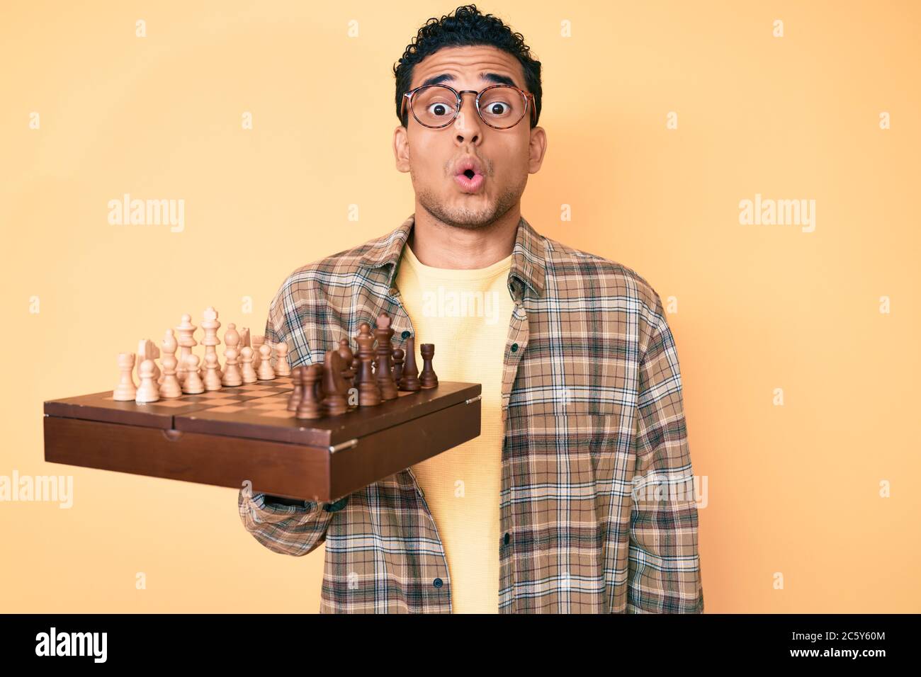 Young handsome hispanic man holding chess board wearing glasses scared ...