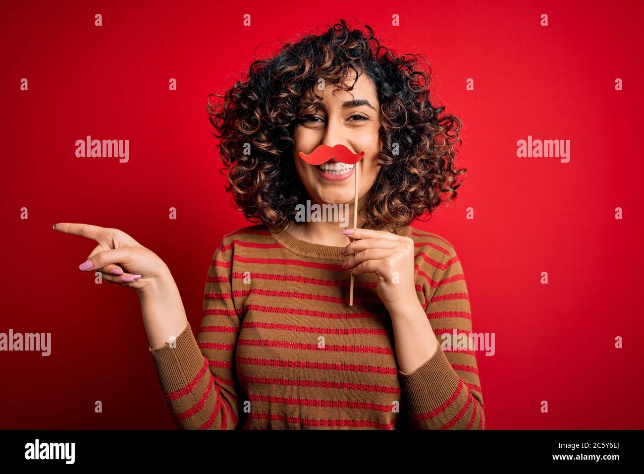 Young beautiful curly arab woman holding funny mustache over isolated ...