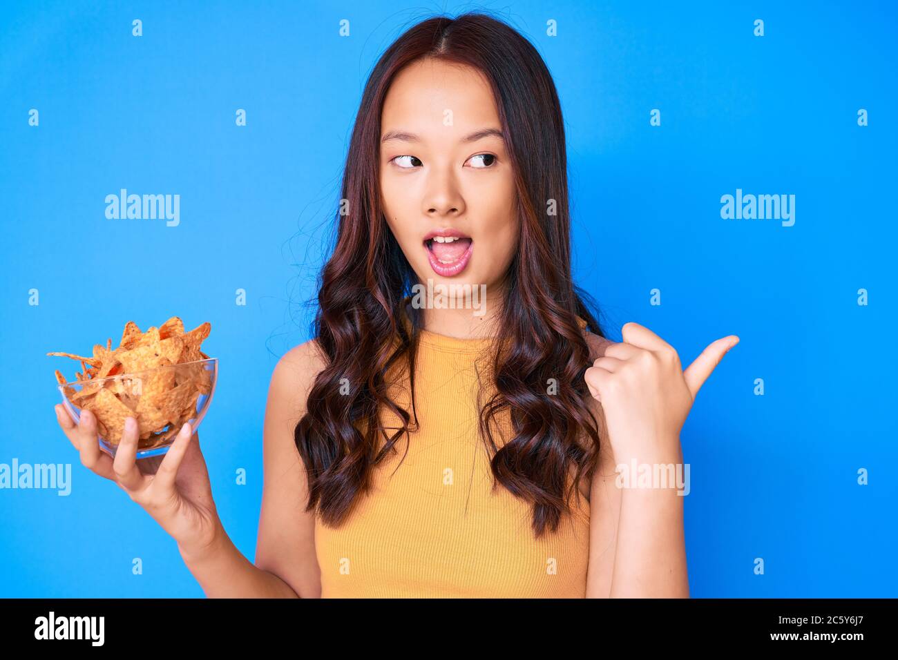 Young beautiful chinese girl holding nachos potato chips pointing thumb ...