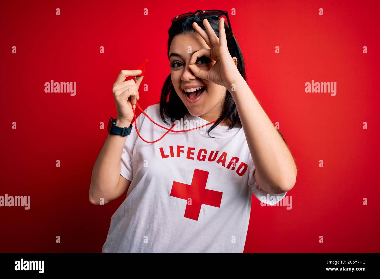 Young lifeguard woman wearing guard equipement holding whistle over red ...