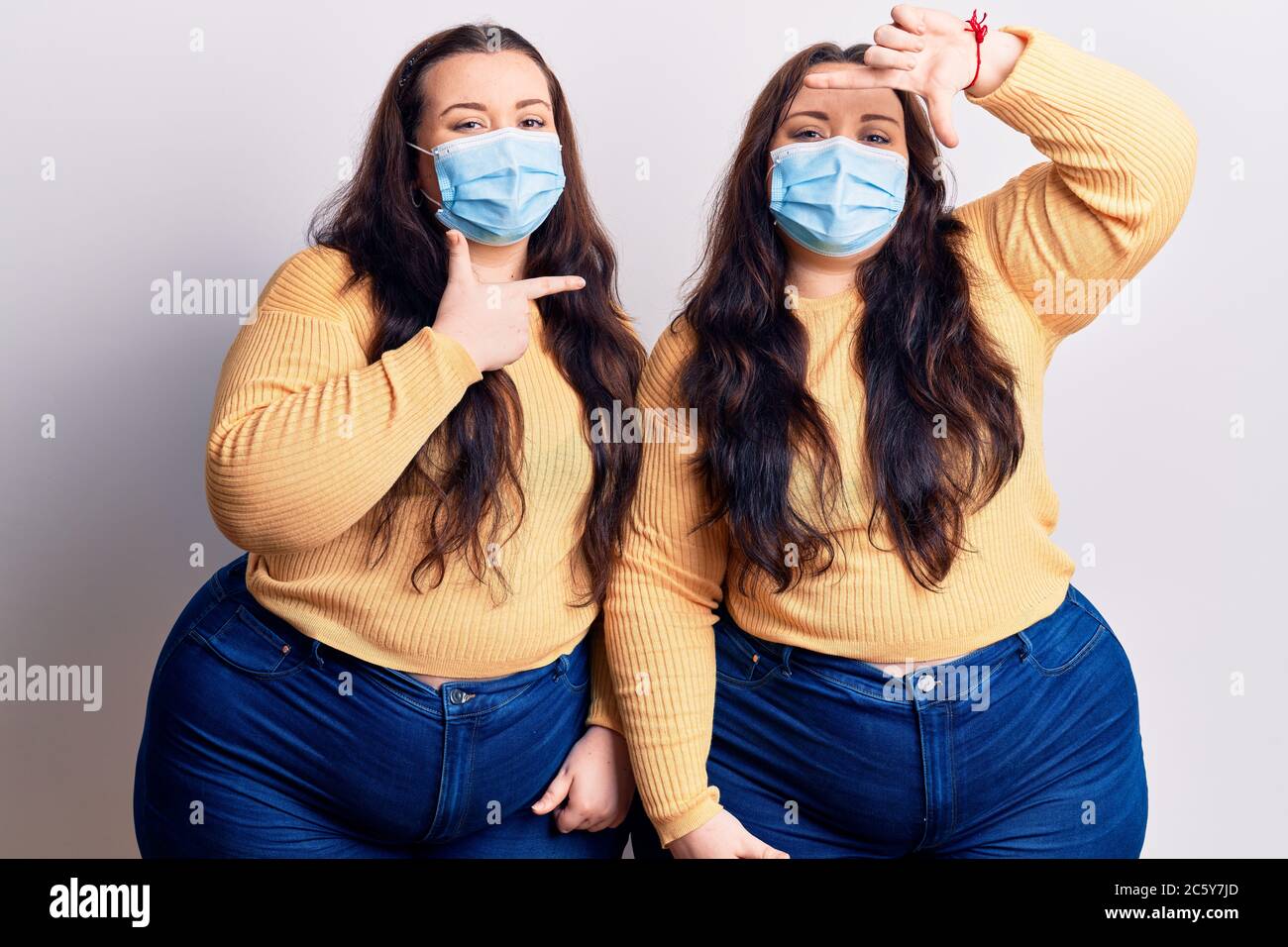 Young plus size twins wearing medical mask smiling making frame with ...