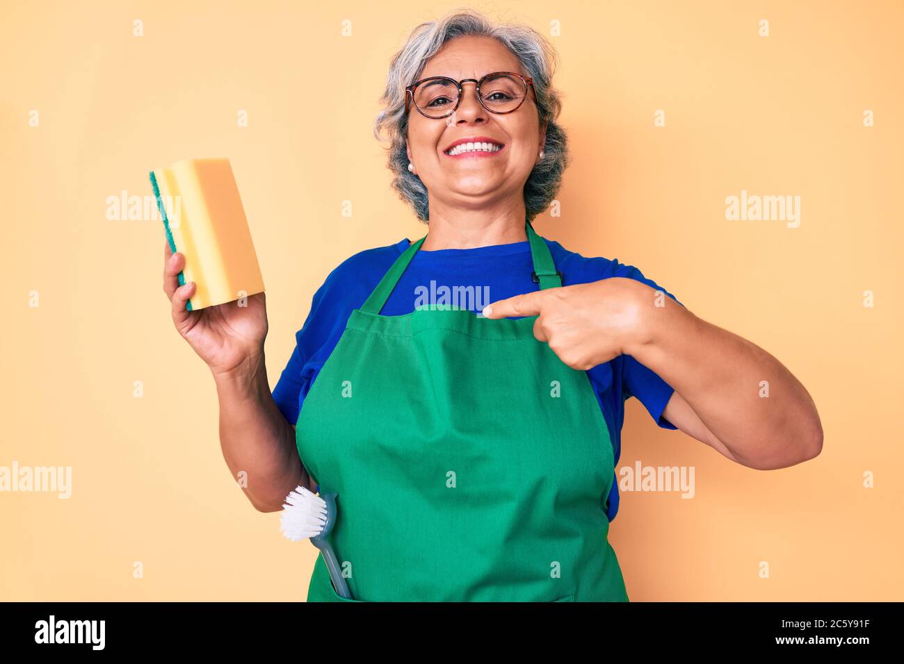 Senior hispanic grey- haired woman wearing apron holding scourer ...
