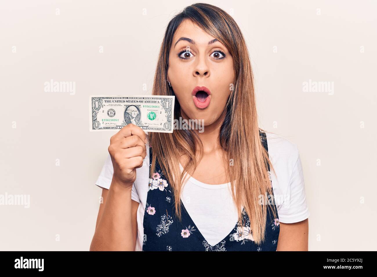 Young beautiful woman holding one dollar scared and amazed with open ...