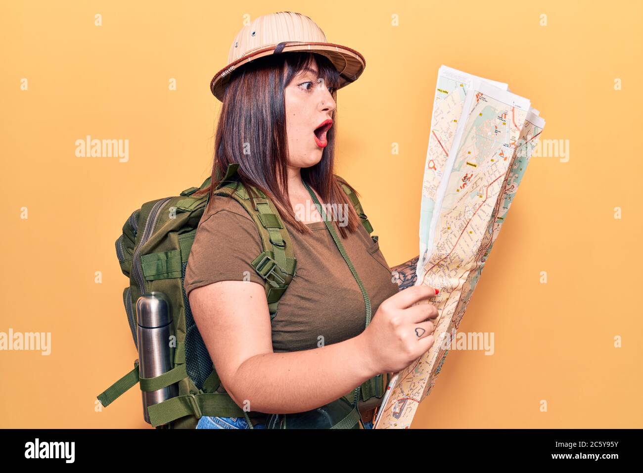 Young plus size woman wearing explorer hat holding map scared and ...