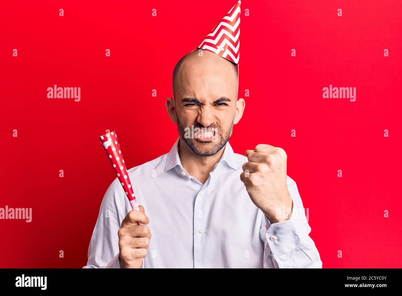 Young handsome bald man wearing party hat and trumpet annoyed and ...