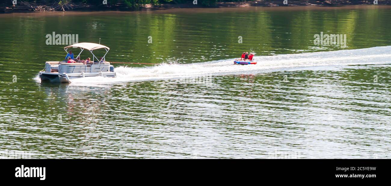 A pontoon boat on the Monongahela River pulling kids on a raft behind ...