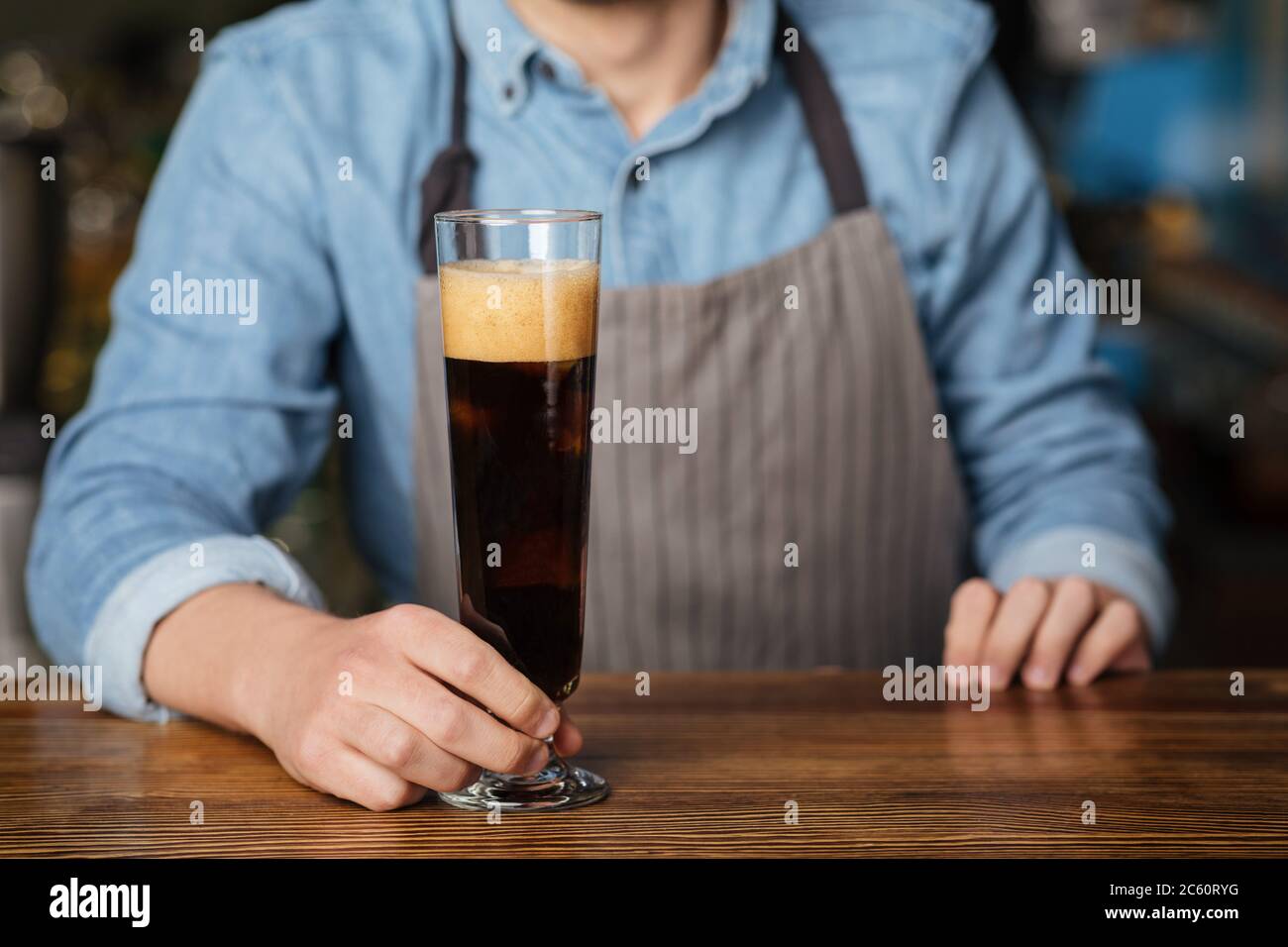 Dark ale with foam. Barman holds high glass of beer on wooden bar ...
