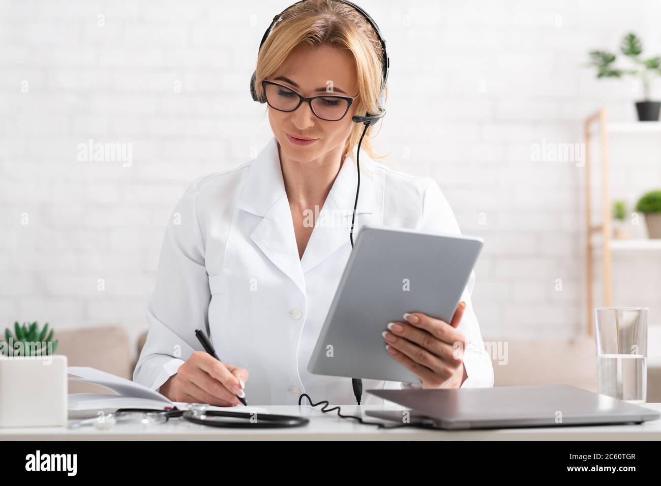 Female doctor makes notes in notebook, holds tablet in hand in interior ...