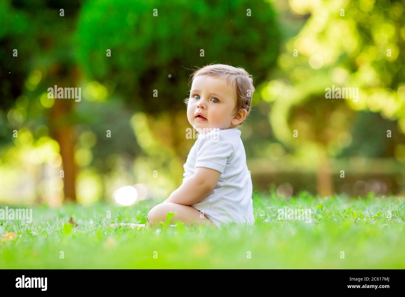 Cute toddler boy of 10 months in white bodysuit sits on green summer ...