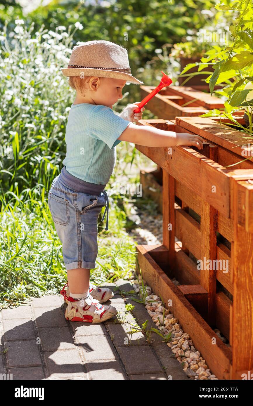 Cute little baby boy wearing lovely hat is gardening by red baby rake ...