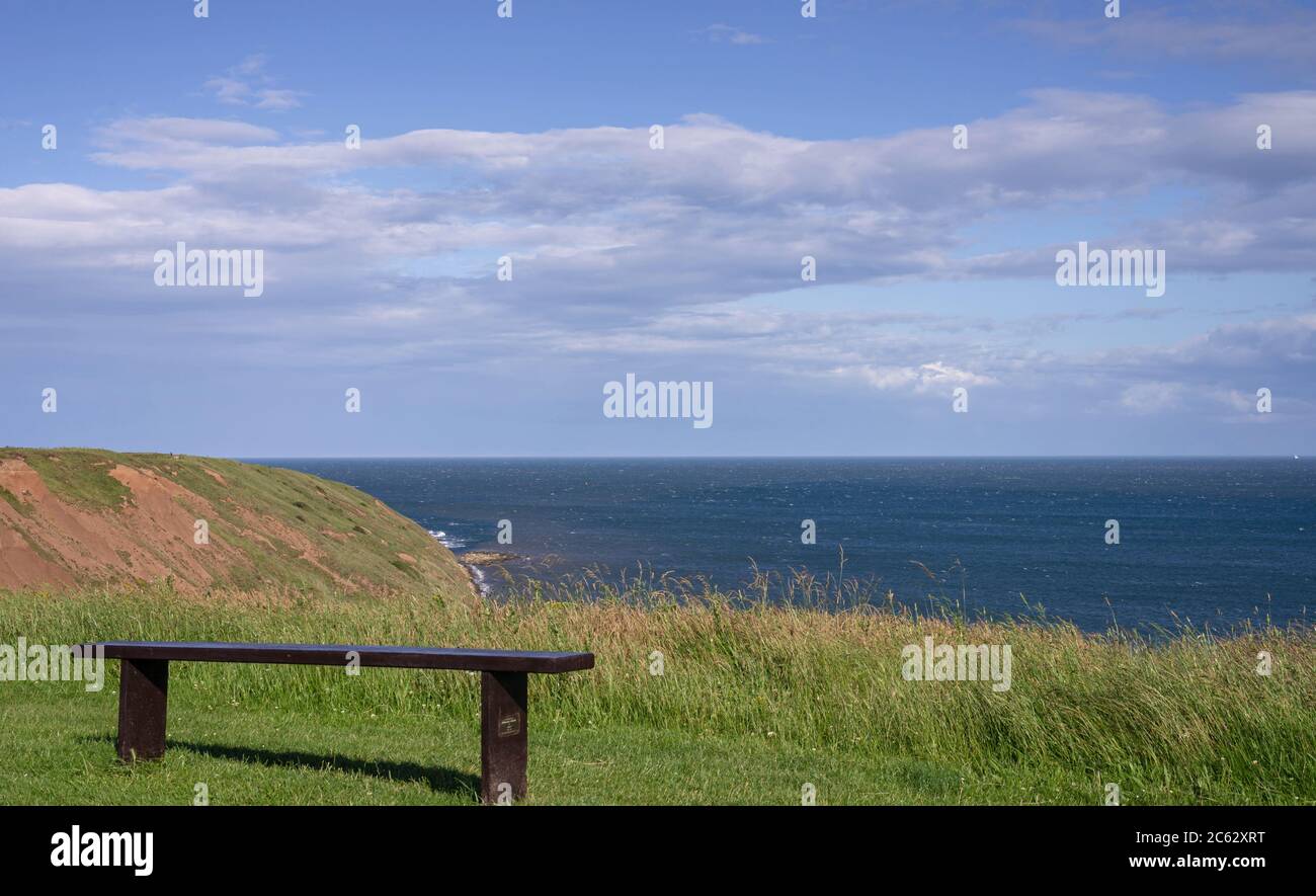 A wooden bench is set on a cliff top looking out to sea. A headland ...