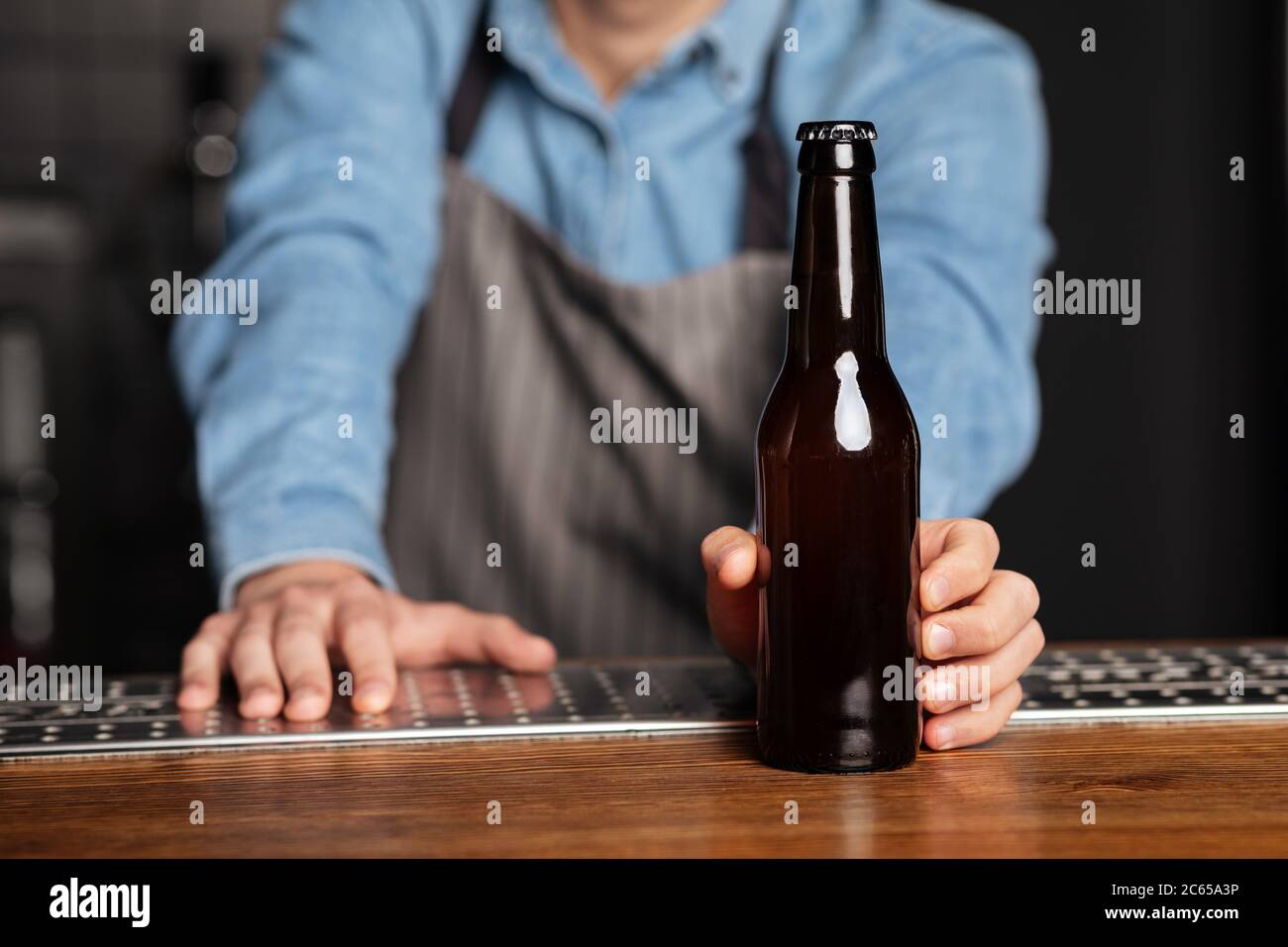 Barman in apron rests on wooden bar counter and serves bottle of beer ...