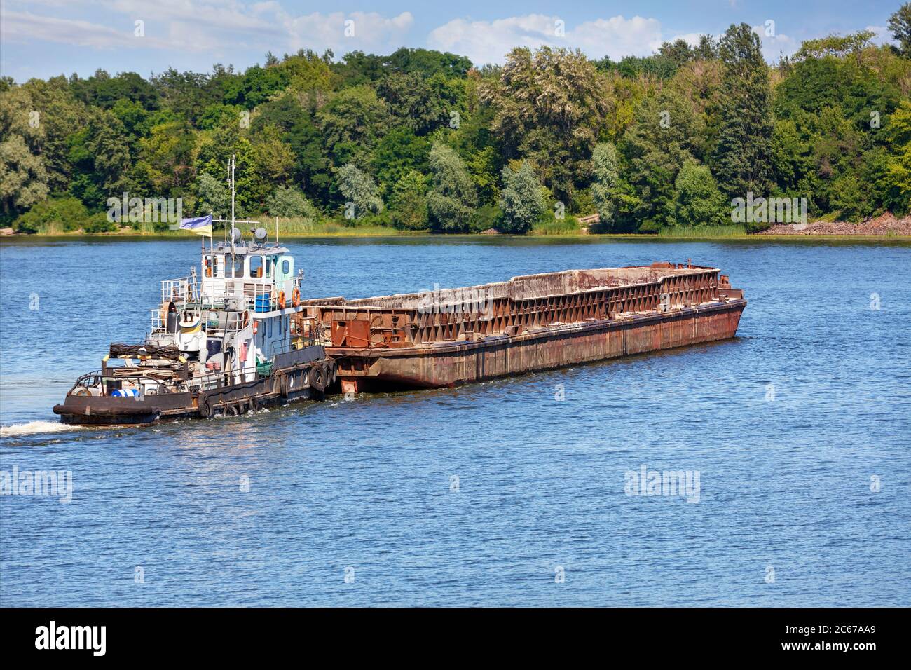 A river tugboat with an empty rusty barge goes crosses a wide river ...