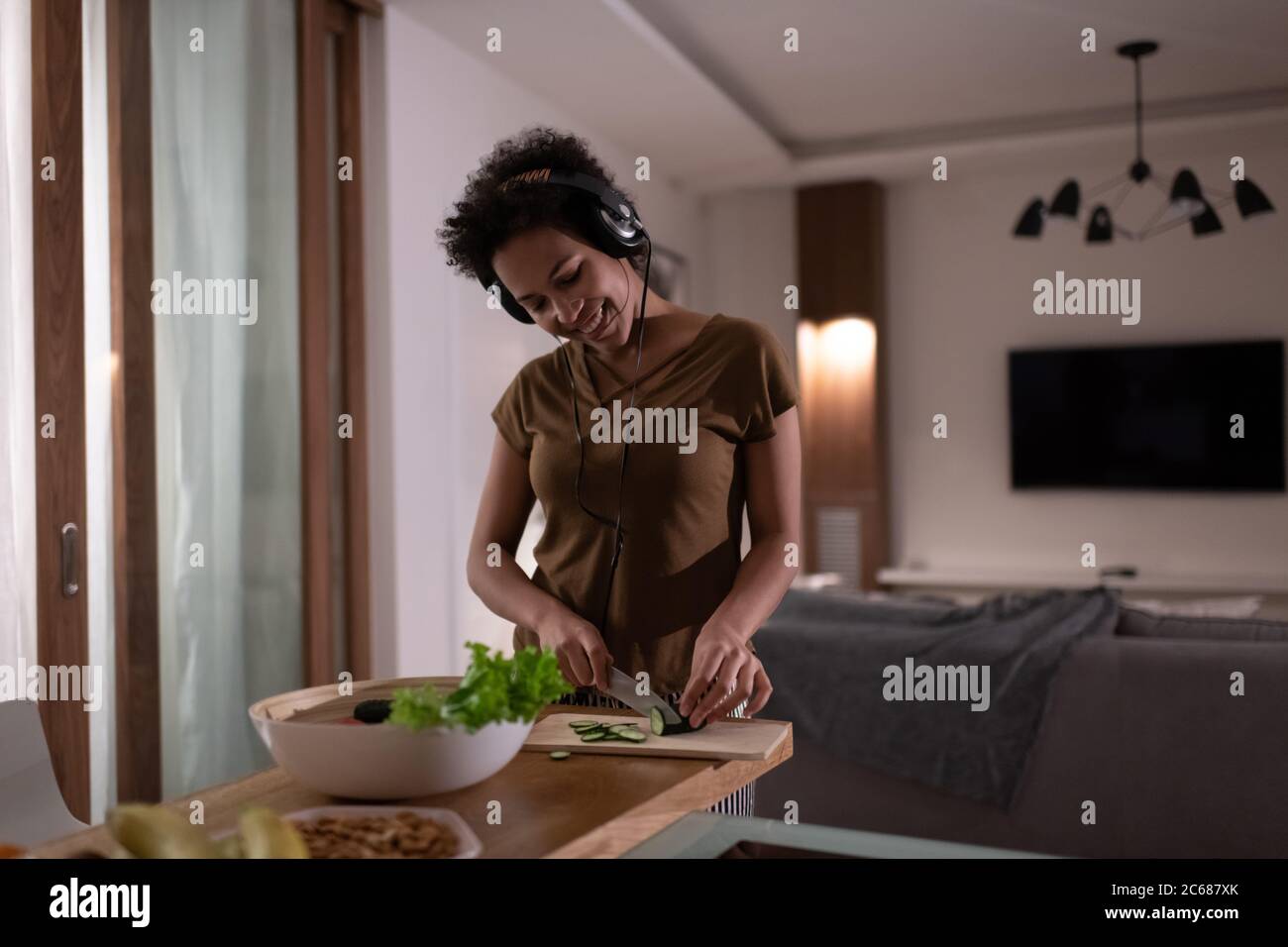 Happy ethnic lady listening to music and cooking healthy salad for ...