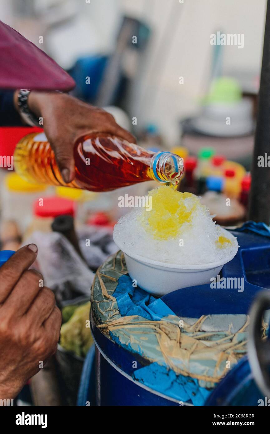 Hand pouring yellow syrup on shaved ice in foam bowl by street vendor ...