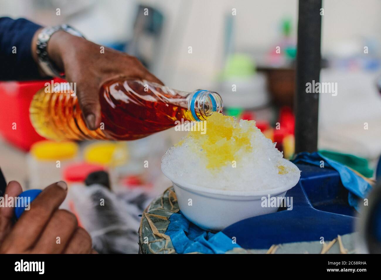 Hand pouring yellow syrup on shaved ice in foam bowl by street vendor ...