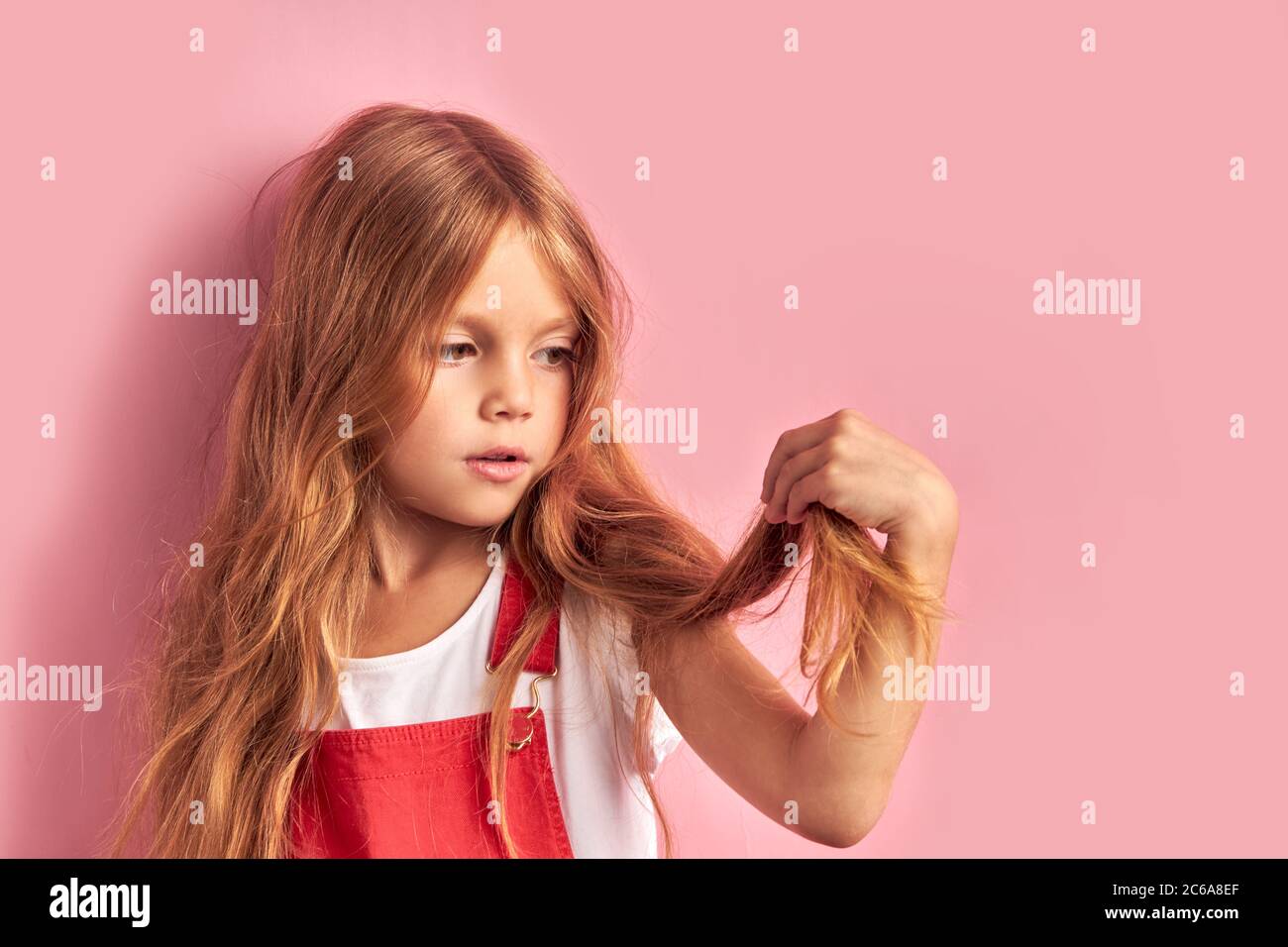 Portrait of worried girl in red overalls examining her hair on head ...