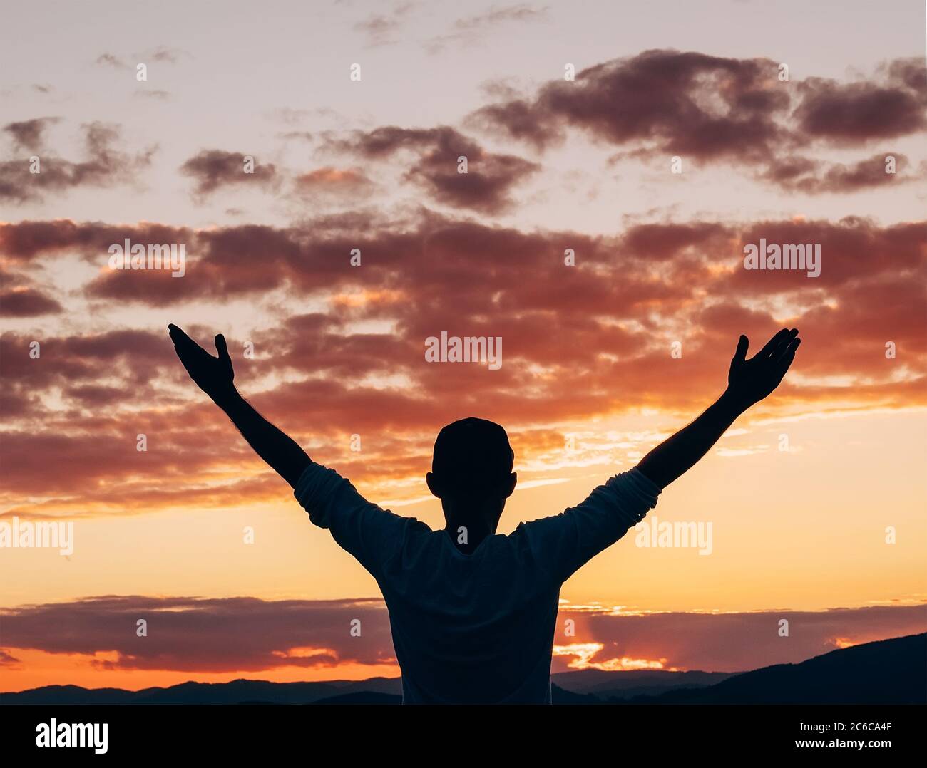 Young man rising up the hands and greeting the stunning sunset orange ...