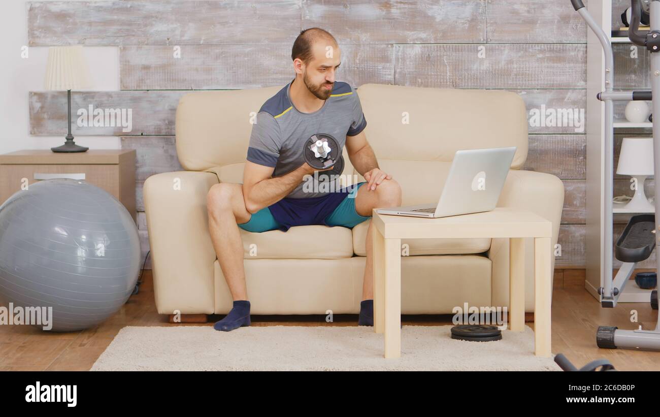 Guy training biceps with dumbbell following online training on laptop ...