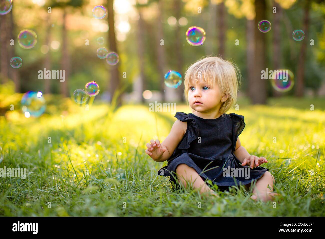 little blonde girl sitting on a grass in a park and playing with soap ...