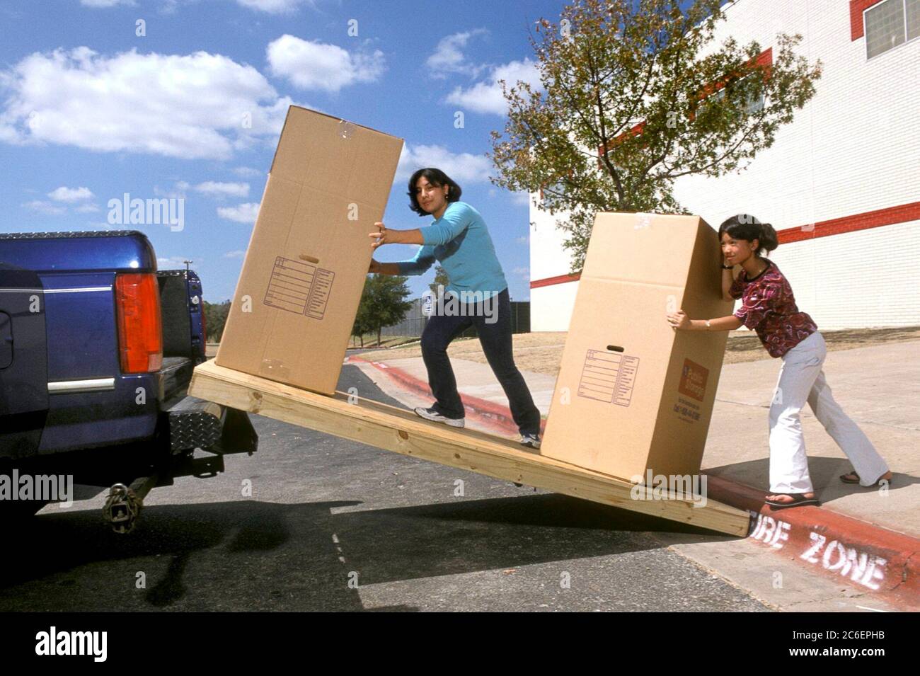 Two 14-year-old Hispanic girls push boxes up inclined plane into back of pickup truck. Model ...