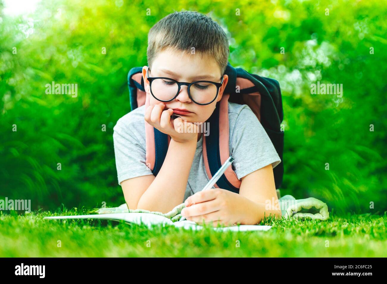 cute schoolboy with backpack in glasses laying on grass making homework ...