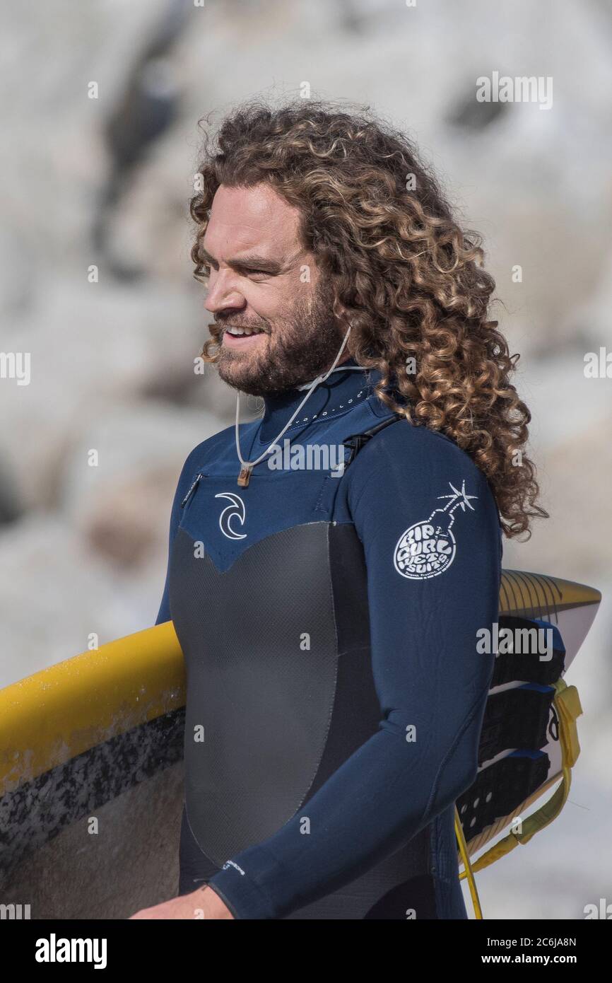A male surfer with long curly hair carrying his surfboard at Fistral in ...