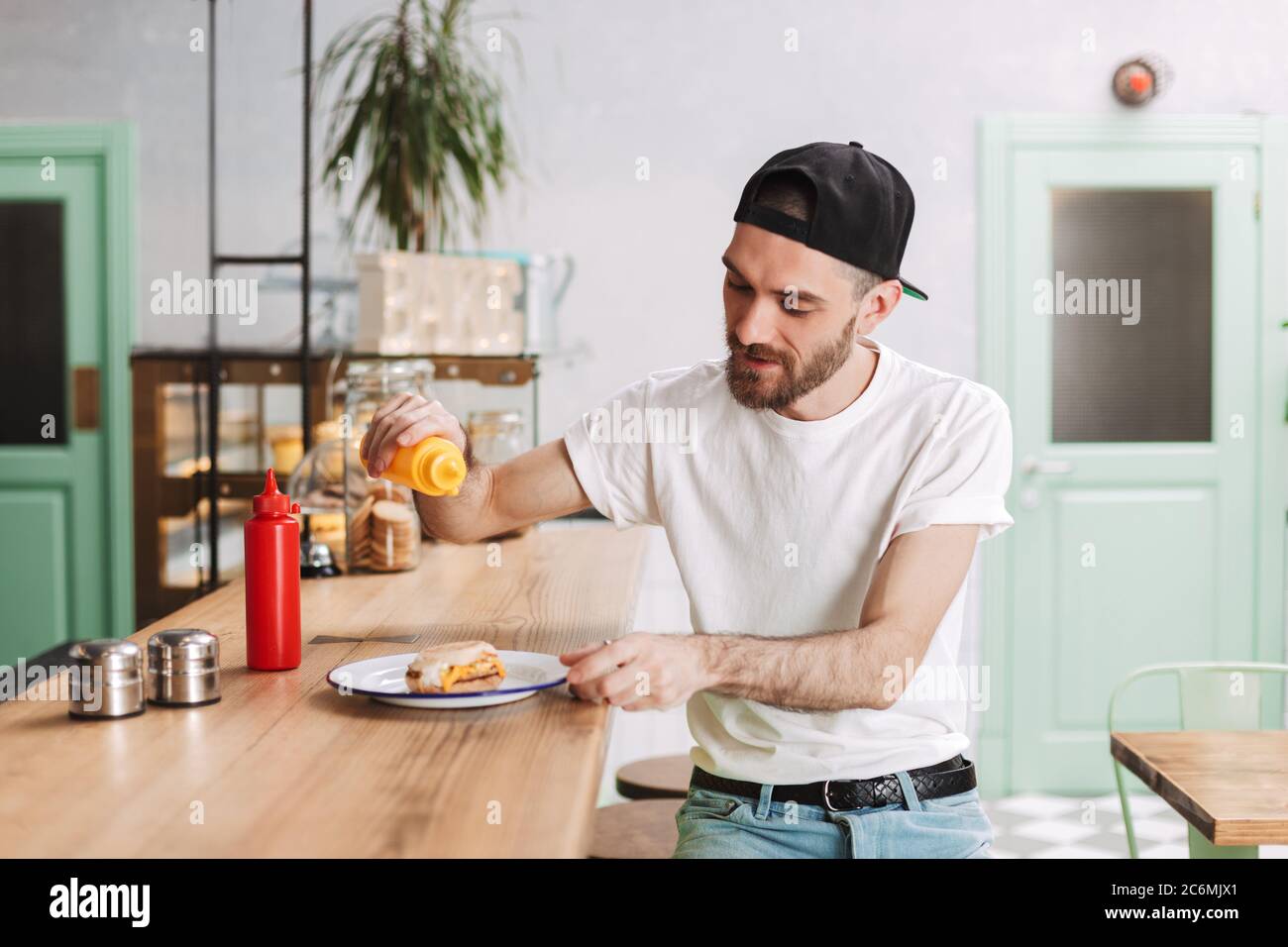 Young man in black cap sitting at the bar counter and holding mustard ...