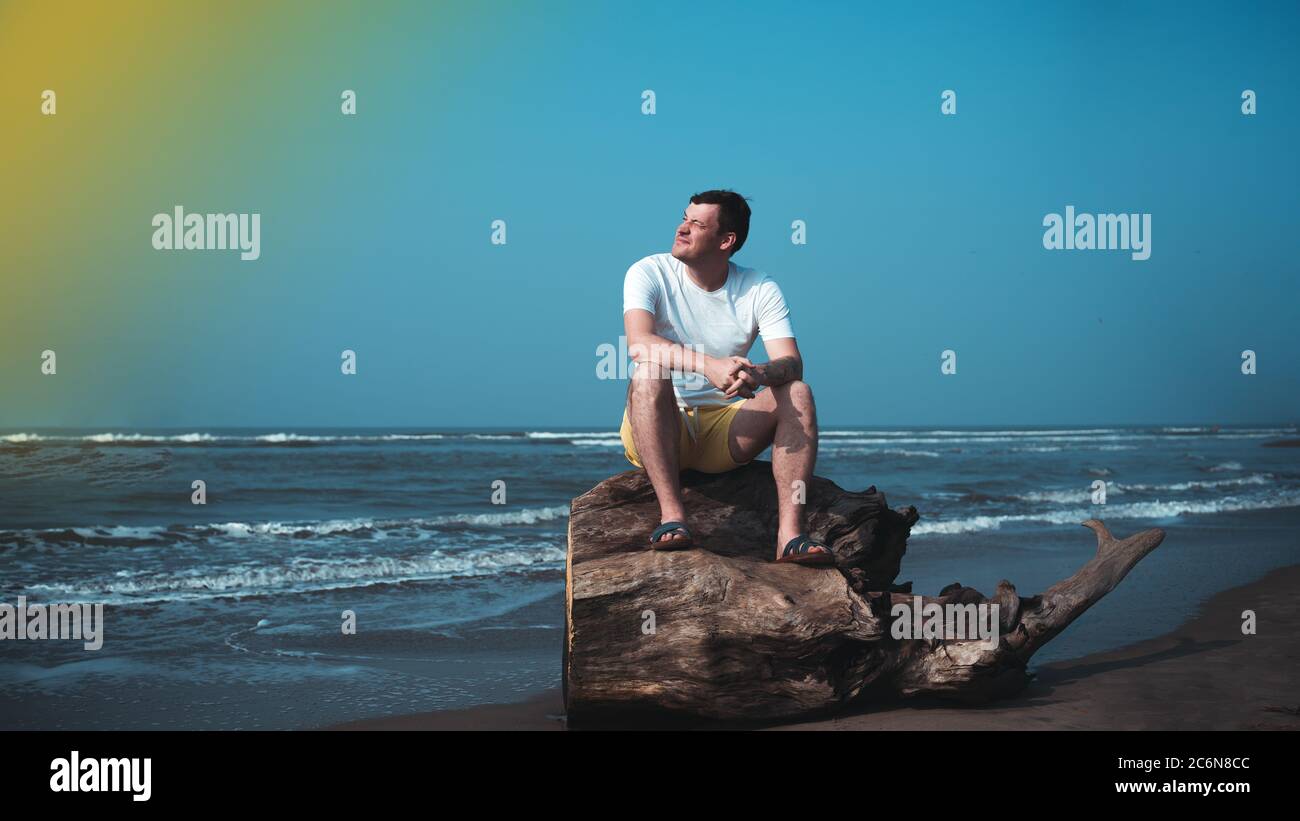 Relaxed man sits on log and looks away. Male tourist sits on driftwood ...