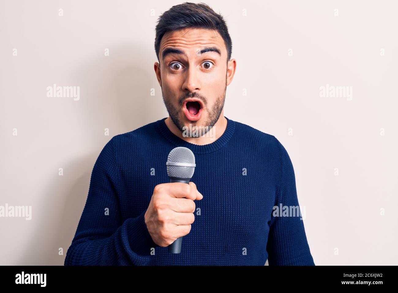 Young handsome singer man with beard singing song using microphone over white background scared ...