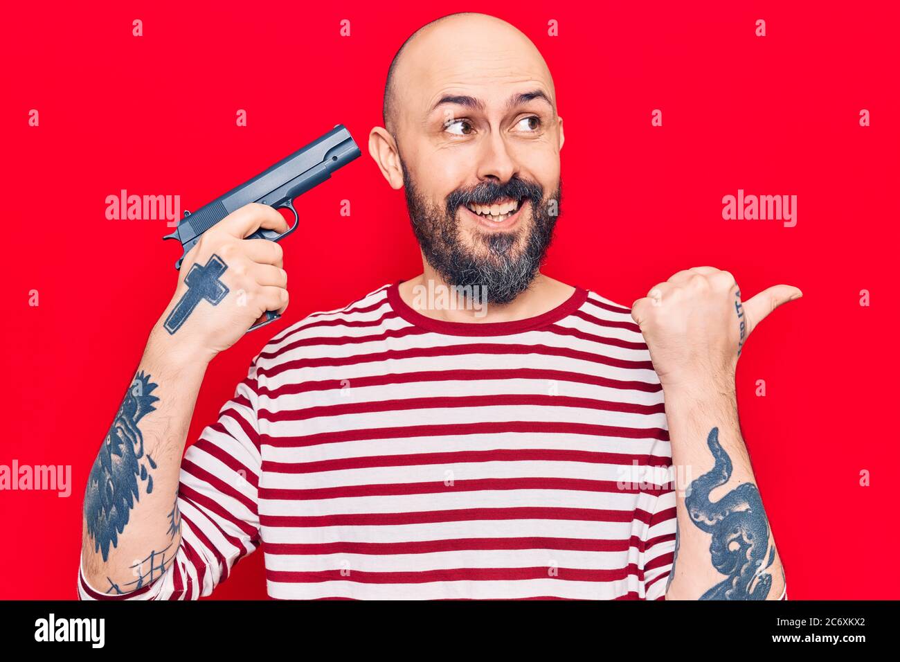 Young handsome man holding gun pointing thumb up to the side smiling ...