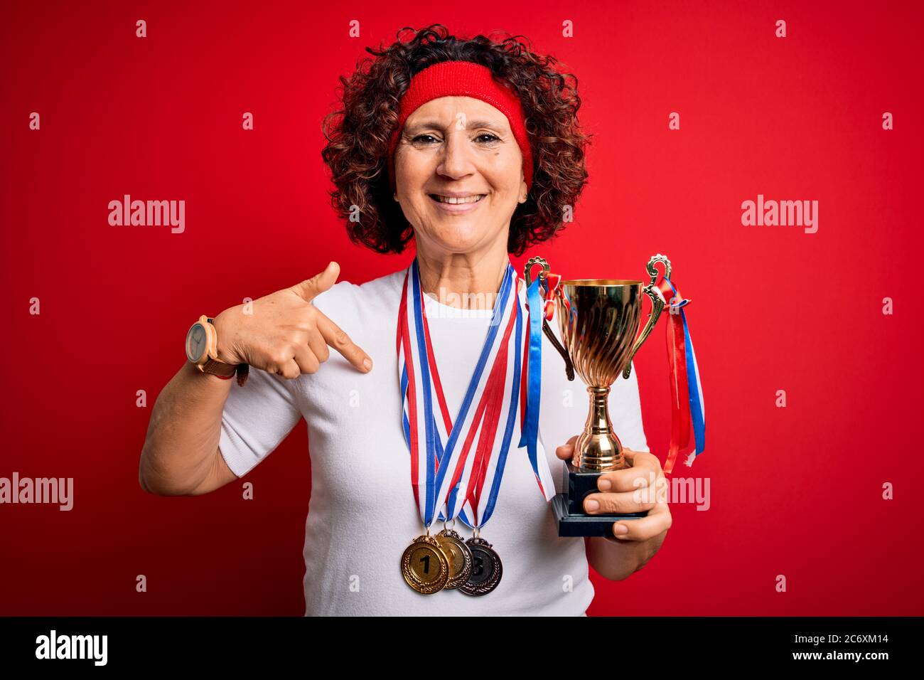 Middle age curly woman winning medals holding trophy over isolated red ...