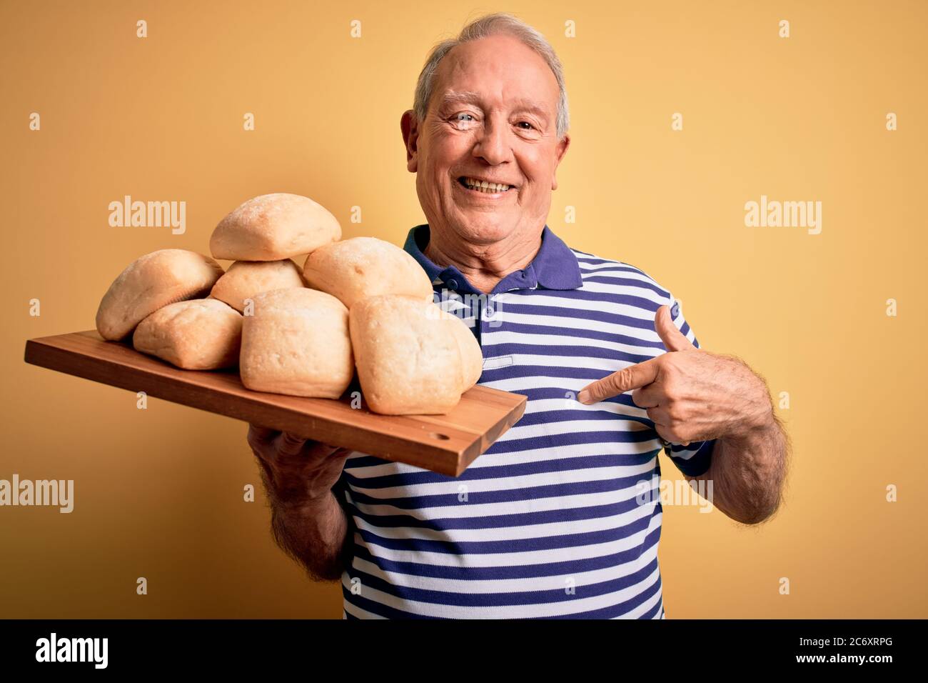 Senior grey haired baker man holding fresh homemade bread over yellow ...