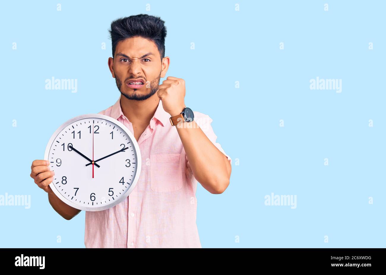 Handsome latin american young man holding big clock annoyed and ...
